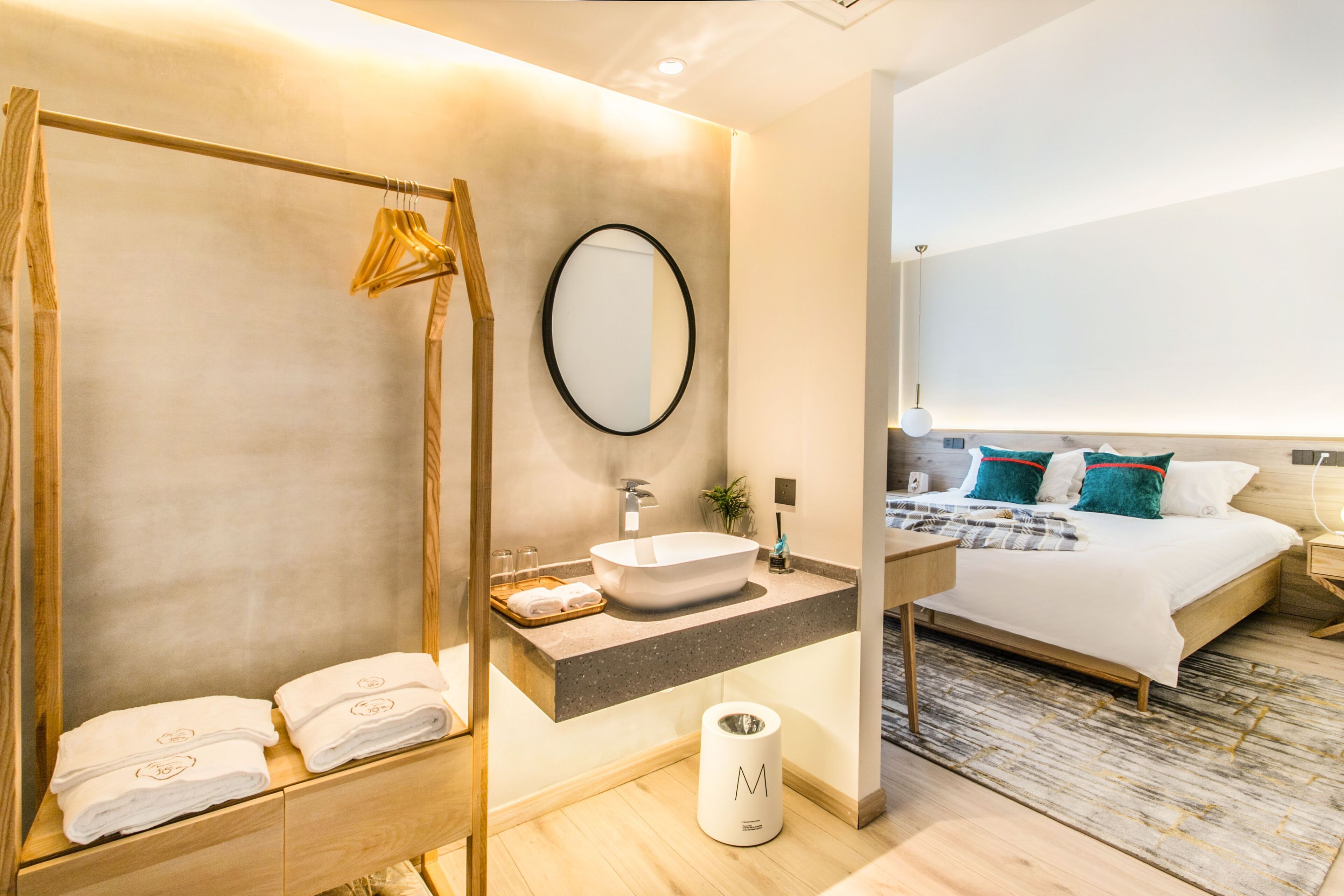 Quarto de hotel moderno com uma cadeira de banho de madeira, um espelho grande, uma pia branca, uma mesa de cabeceira, uma cama com cabeceira cinza e um tapete estampado.