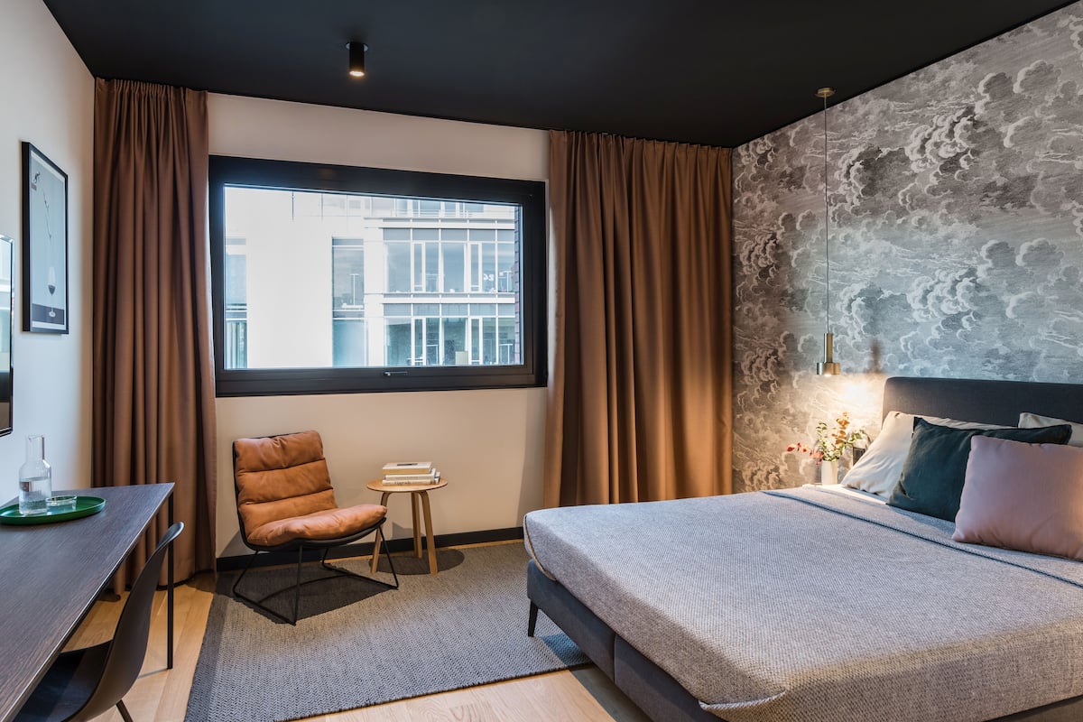 Quarto de hotel moderno com uma cama grande, uma escrivaninha com cadeira, uma pequena mesa de apoio e vista para a cidade através de uma janela ampla.