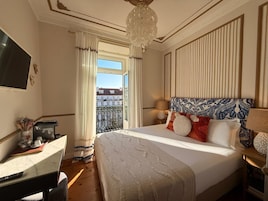 Quarto de hotel com uma cama grande, uma escrivaninha com cadeira, televisão e varanda com cortinas.