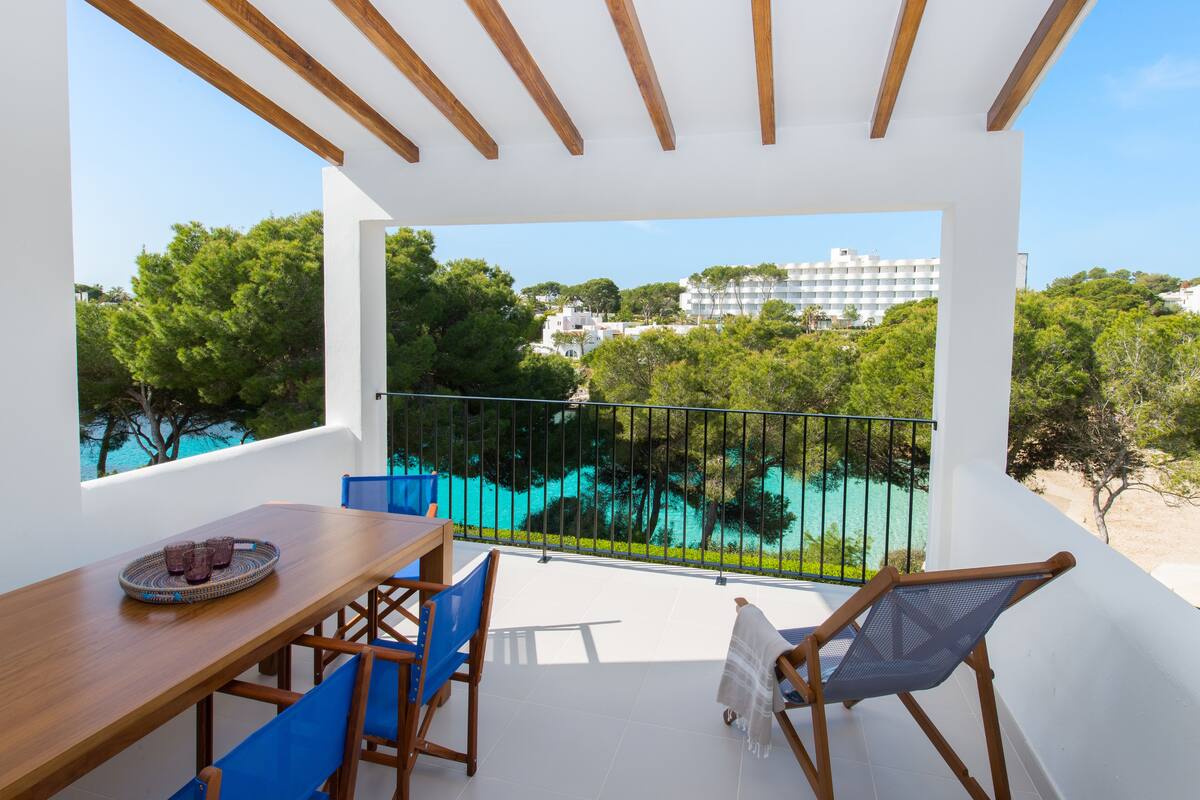 Uma varanda com mesa de madeira, cadeiras azuis e vista para uma piscina e edifícios.