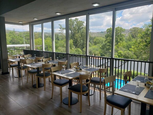 Uma área de refeições com mesas e cadeiras, grandes janelas com vista para vegetação e colinas.