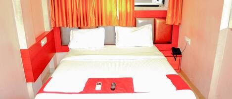 Quarto de hotel com cama, mesa de cabeceira, esquema de cores vermelho e branco, e janela com cortinas.
