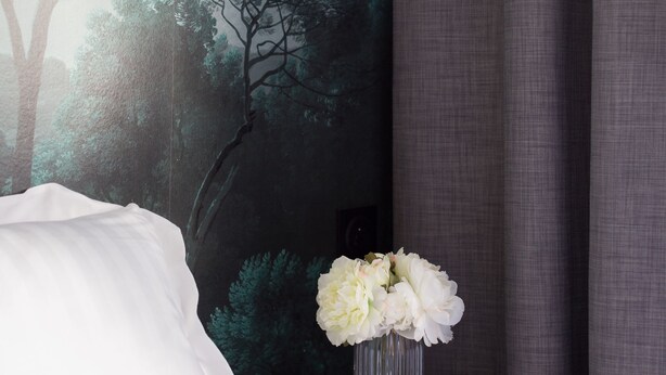 Quarto com cama, um vaso de flores, luminária na parede e pintura de árvores.