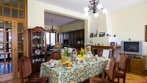 Uma sala de jantar com uma mesa posta para quatro pessoas, rodeada por cadeiras de madeira. Um candelabro pendurado acima e uma televisão visível ao fundo.