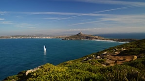 Paisagem costeira com um farol numa colina, um veleiro na água e uma linha de costa distante com edifícios.
