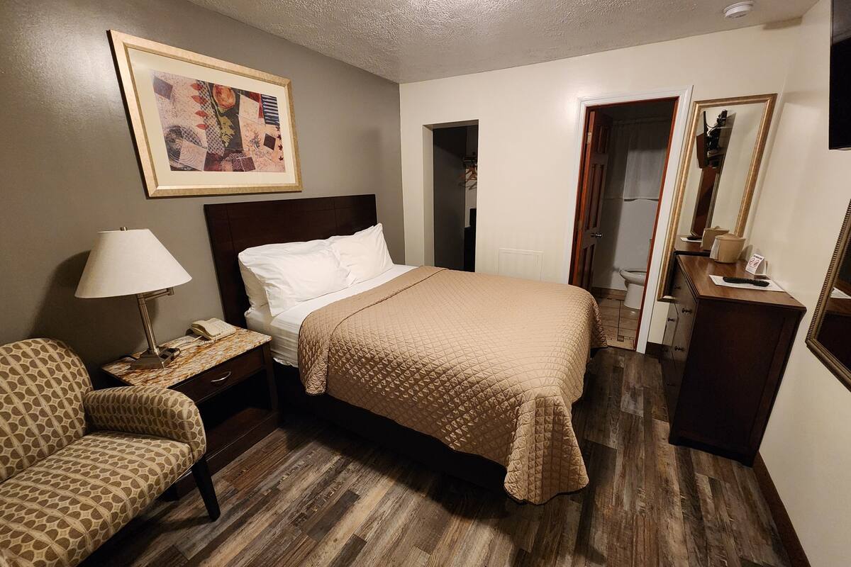 Quarto de hotel com cama, cadeira, escrivaninha com luminária, espelho e quadro na parede.