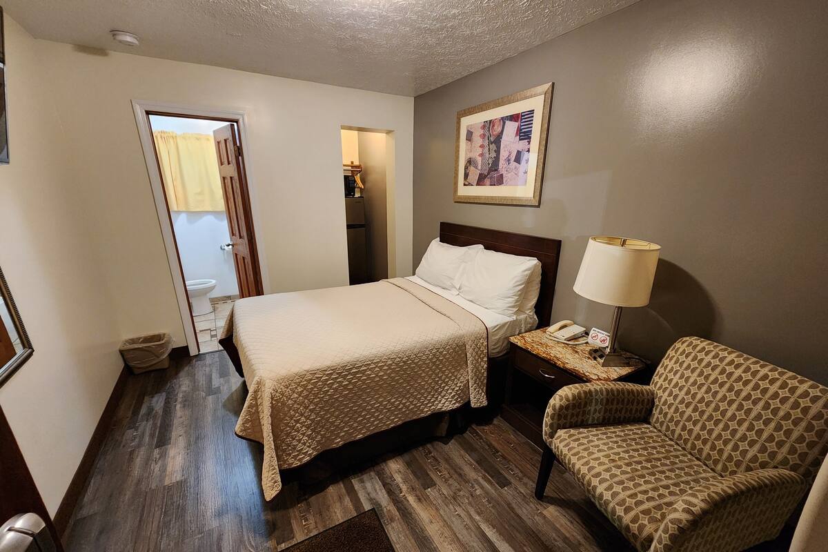 Quarto de hotel com cama, uma cadeira, mesa de cabeceira com abajur, quadro na parede e porta para o banheiro.