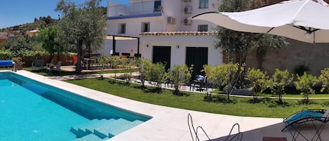 Piscina com design de azulejos azuis, rodeada por um espaço gramado com espreguiçadeiras e um guarda-sol branco.