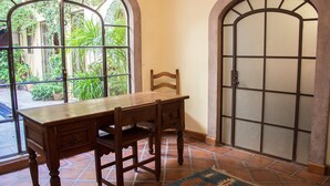 Uma escrivaninha de madeira com uma cadeira em um quarto com chão de azulejos e uma janela grande com grades de metal.