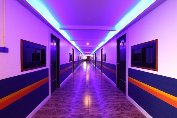 Corredor comprido e estreito, com portas em ambos os lados, iluminado por luzes roxas e azuis.