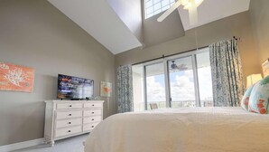 Quarto com uma cama grande, televisão sobre uma cômoda, janela com cortinas e um ventilador de teto.