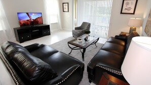 Uma sala de estar com sofá de couro preto, poltrona estampada, mesa de centro de vidro, televisão sobre um suporte preto e uma janela grande com persianas.