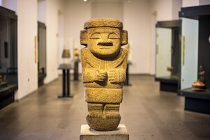 Escultura de pedra em corredor de museu, com piso polido e vitrines de vidro.