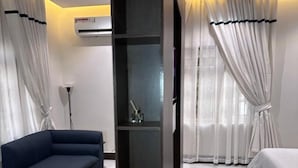 Quarto de hotel moderno com sofá, cama, tapete grande e uma estante alta e escura.
