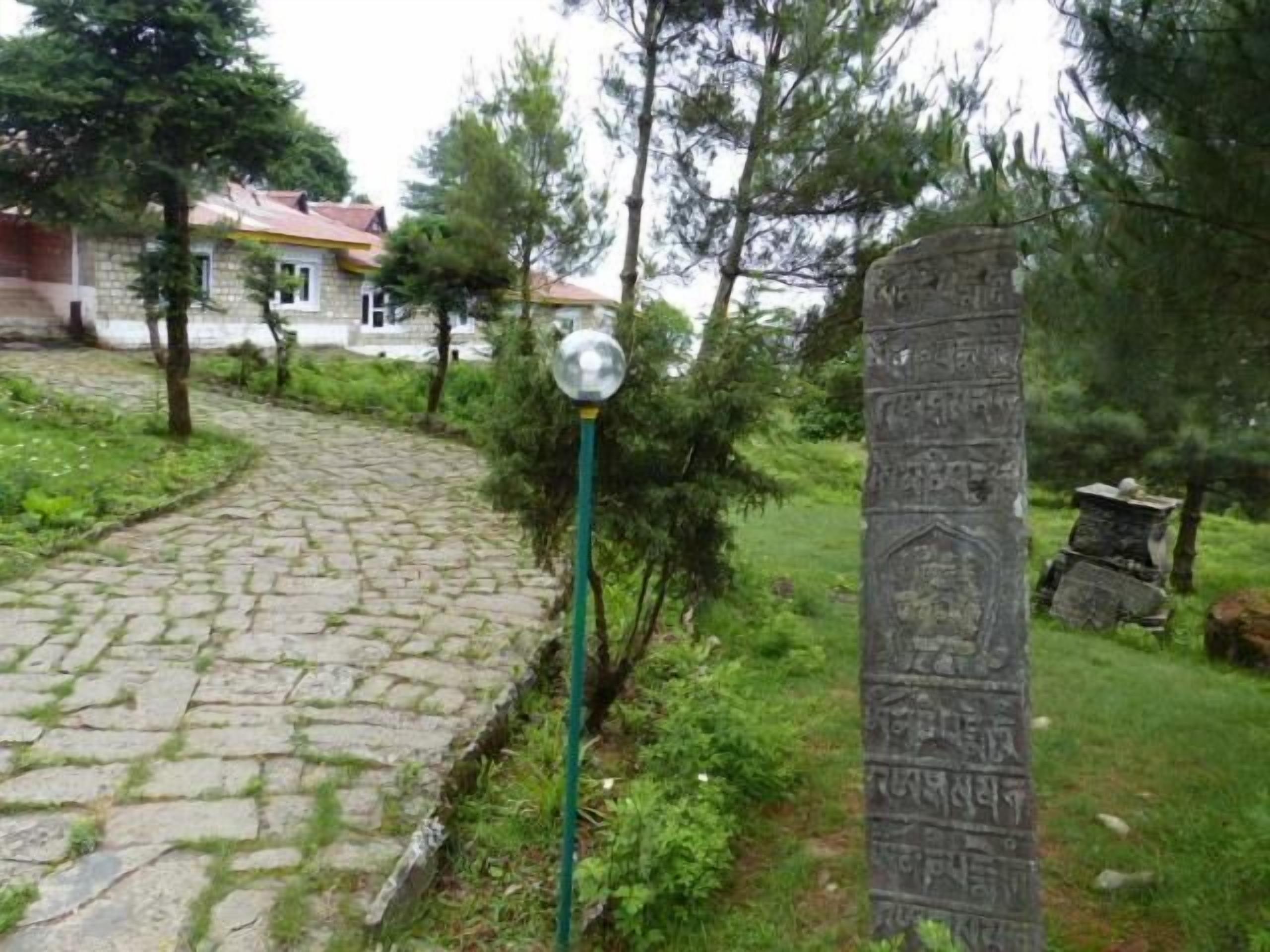 Uma trilha de pedra leva a um monumento de pedra com inscrições, rodeado por vegetação e um edifício com telhado vermelho ao fundo.