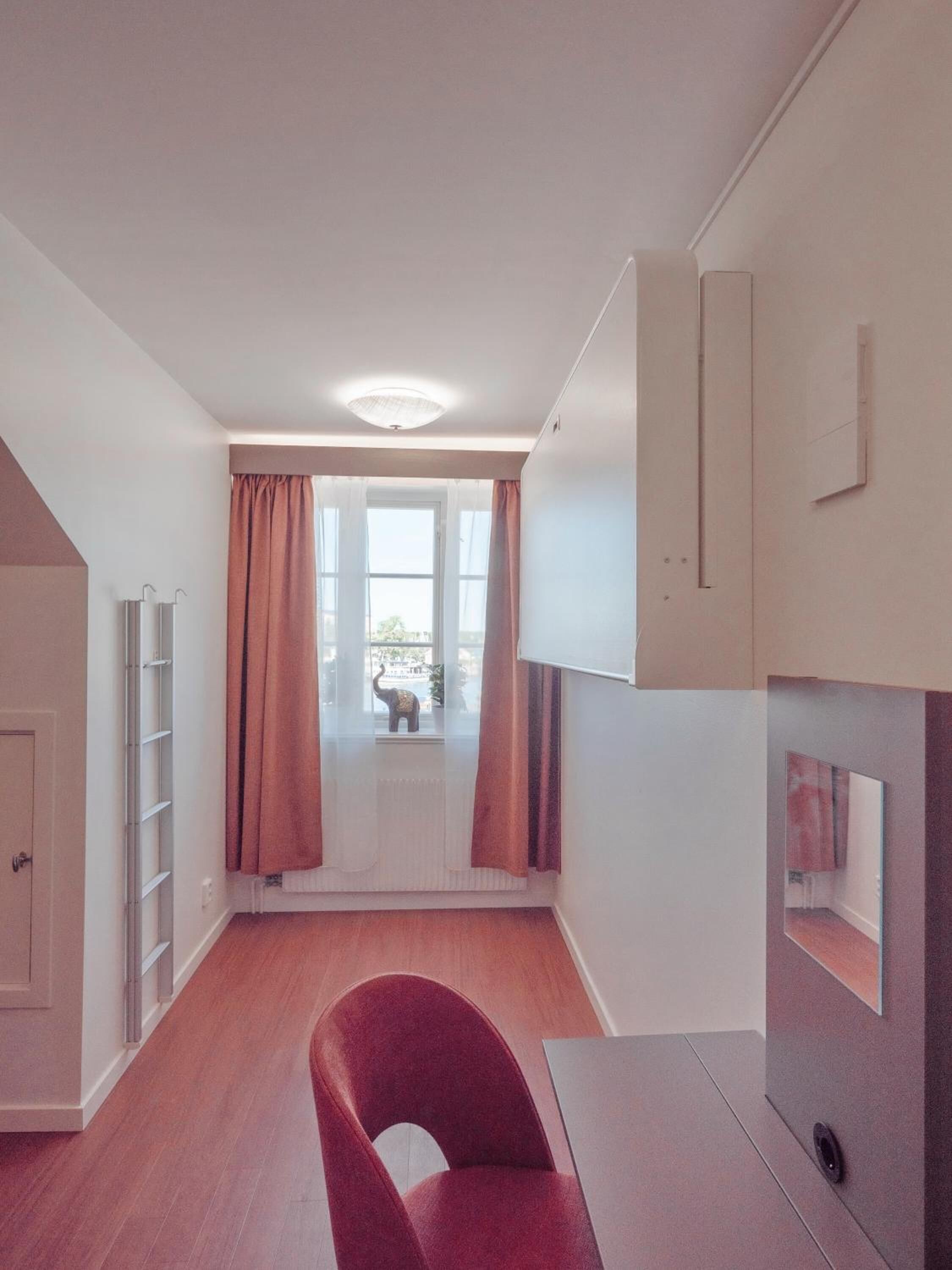 Um corredor estreito com uma cadeira vermelha, uma porta e uma janela com cortinas.