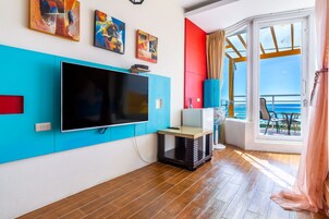 Uma sala de estar moderna com uma grande televisão de tela plana, obras de arte vibrantes e vista para o mar através de uma porta de vidro deslizante.