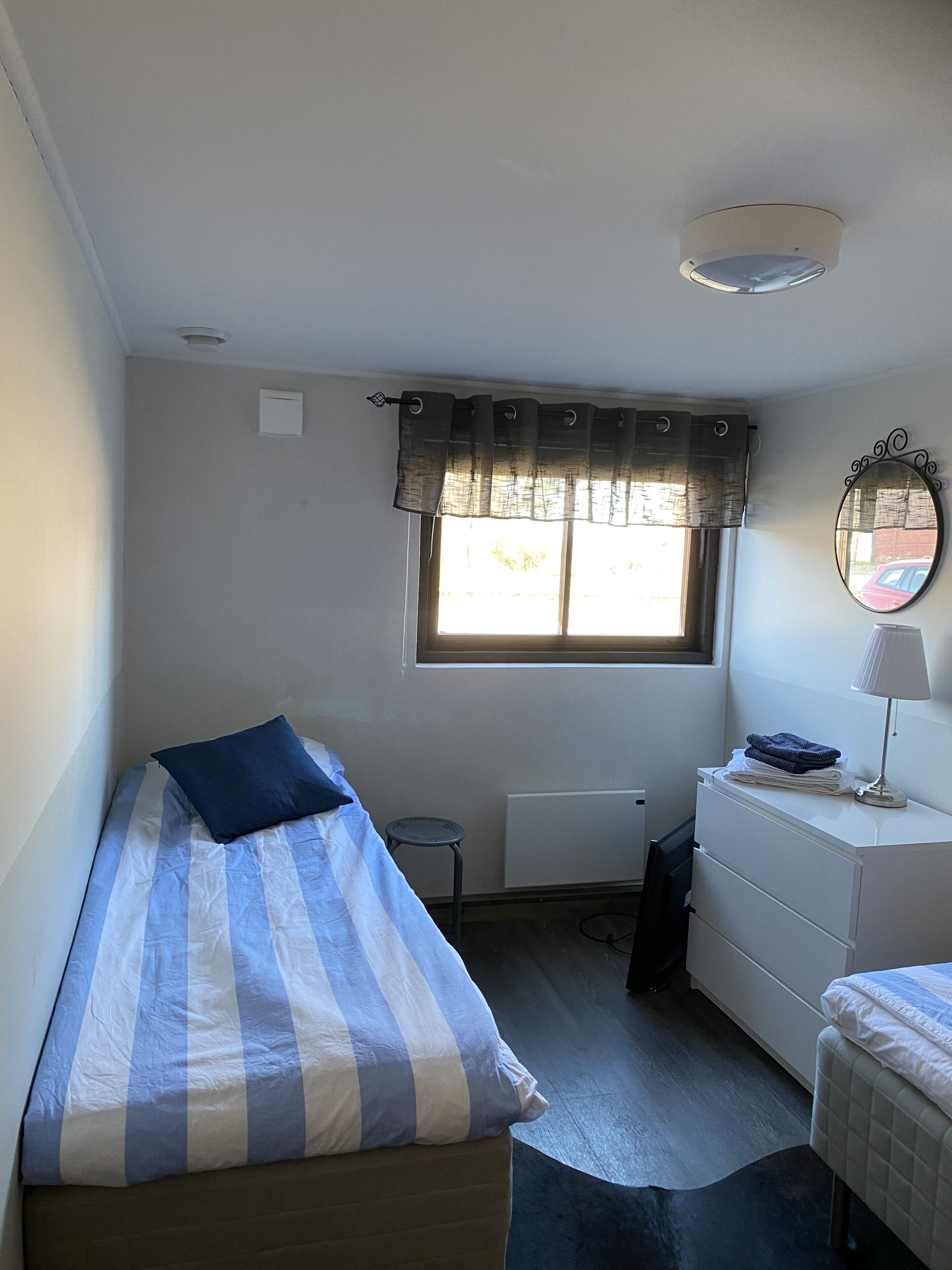 Quarto pequeno e bem arrumado, com cama, penteadeira, espelho e janela com cortinas.