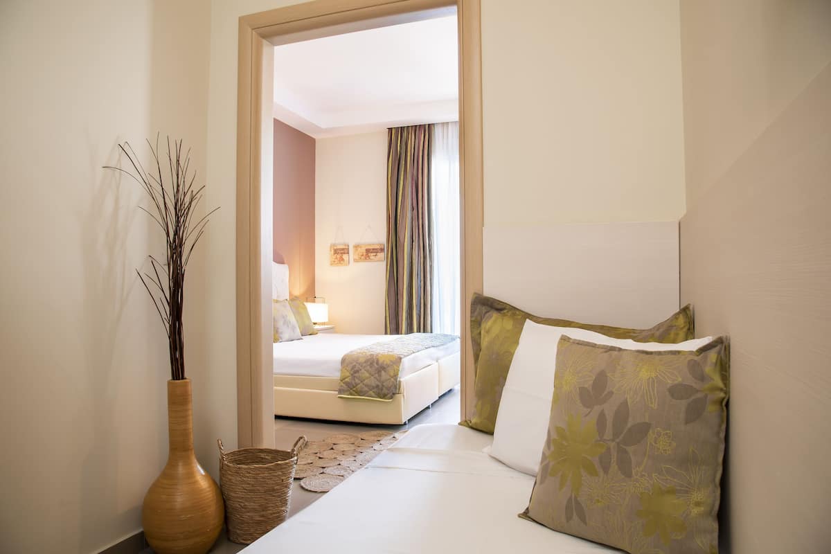 Quarto de hotel com uma cama, um vaso com ramos, uma cesta de vime e uma almofada estampada com flores.