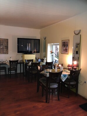 Uma área de refeições com uma TV na parede, uma mesa de jantar posta com cadeiras e uma janela com vista para um edifício.