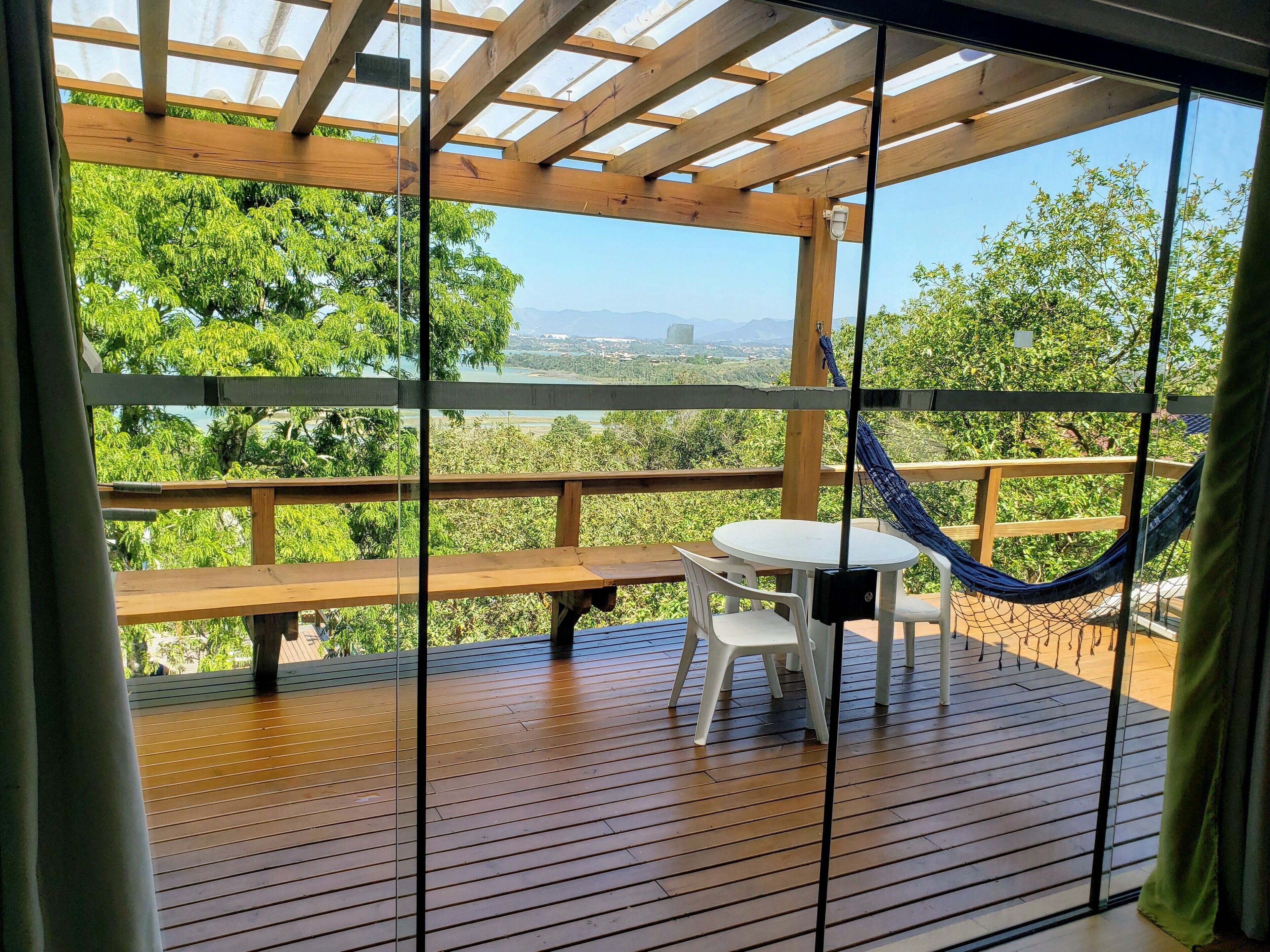 Um deck de madeira com mesa, cadeiras, uma rede e vista para vegetação e montanhas ao longe.