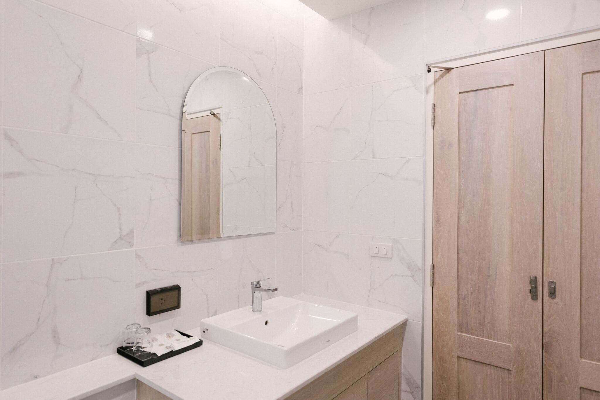 Casa de banho moderna com painel de mármore branco, um espelho grande, lavatório branco e portas de madeira.