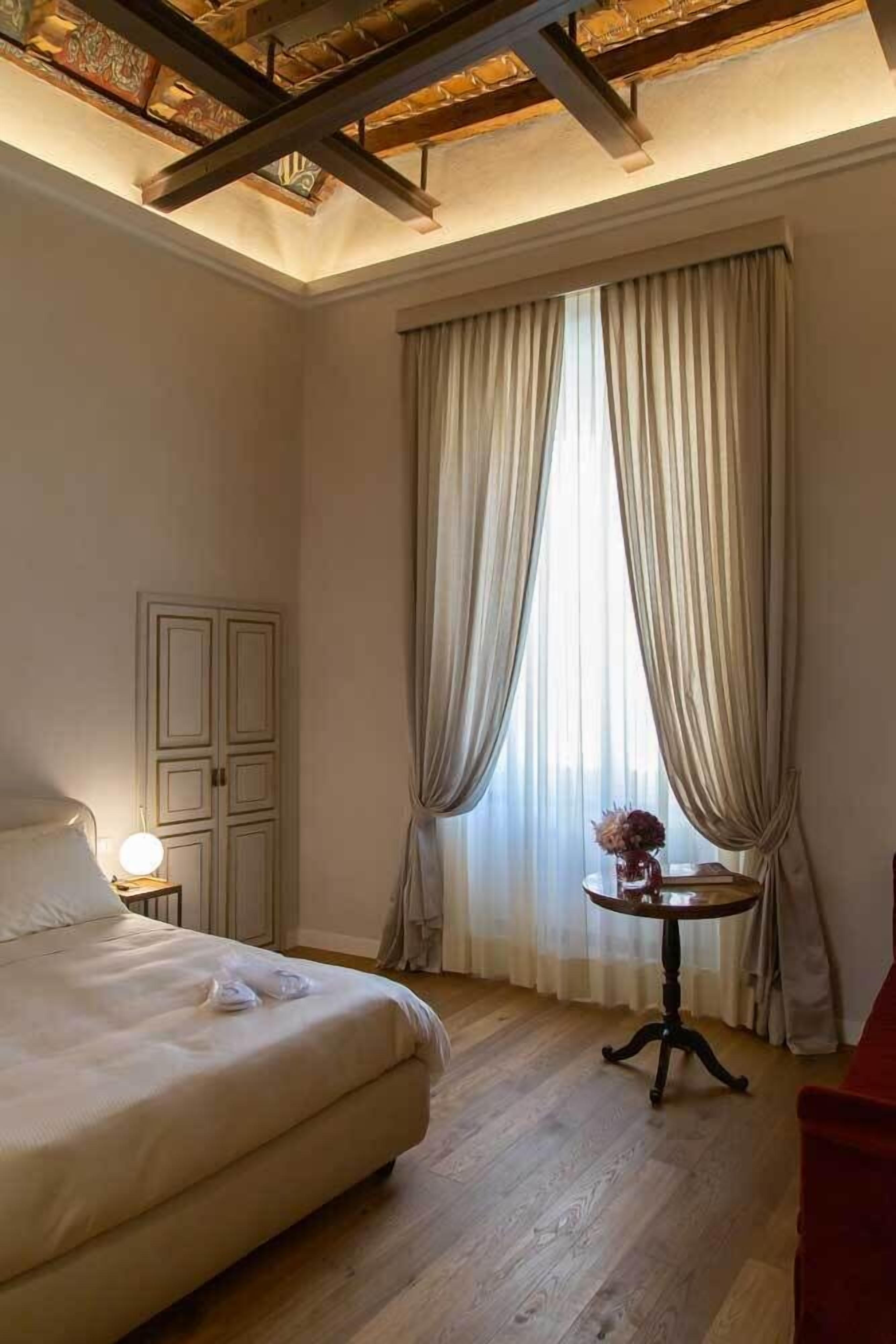Quarto com cama, mesa de cabeceira, mesa com um vaso e janela com cortinas.