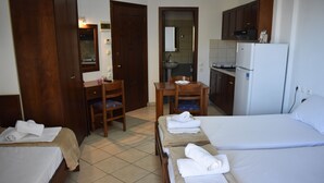 Quarto de hotel com duas camas, uma escrivaninha, uma cadeira, uma pequena cozinha e um banheiro visível por uma porta aberta.