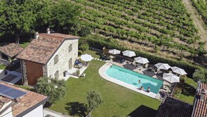 Uma villa com piscina, rodeada por guarda-sóis, um edifício de pedra e um vinhedo ao fundo.