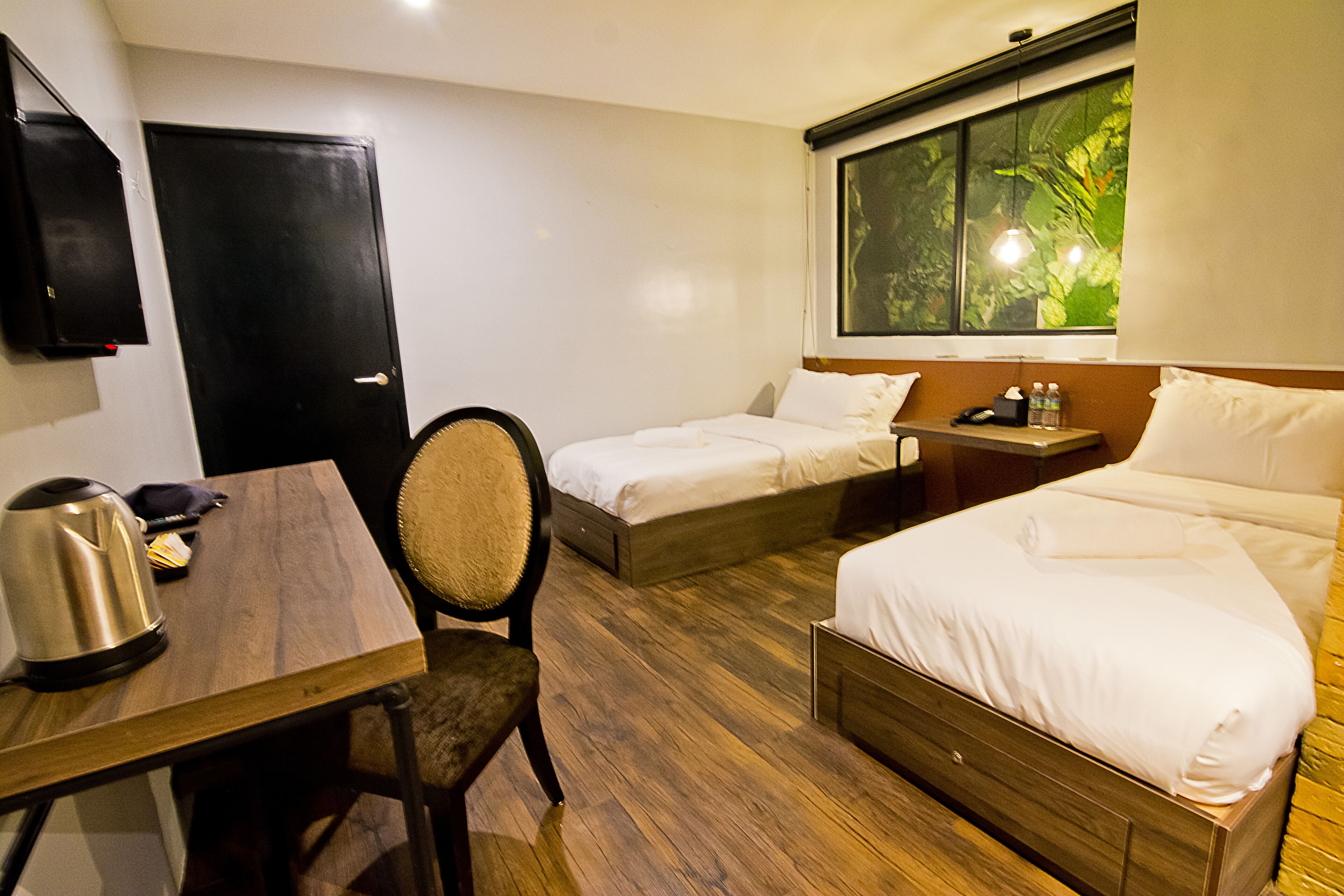Quarto de hotel com piso de madeira, duas camas de solteiro, uma escrivaninha com chaleira, uma cadeira, uma TV e uma janela com vista para área verde.