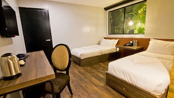 Quarto de hotel com piso de madeira, duas camas de solteiro, uma escrivaninha com chaleira, uma cadeira, uma TV e uma janela com vista para área verde.