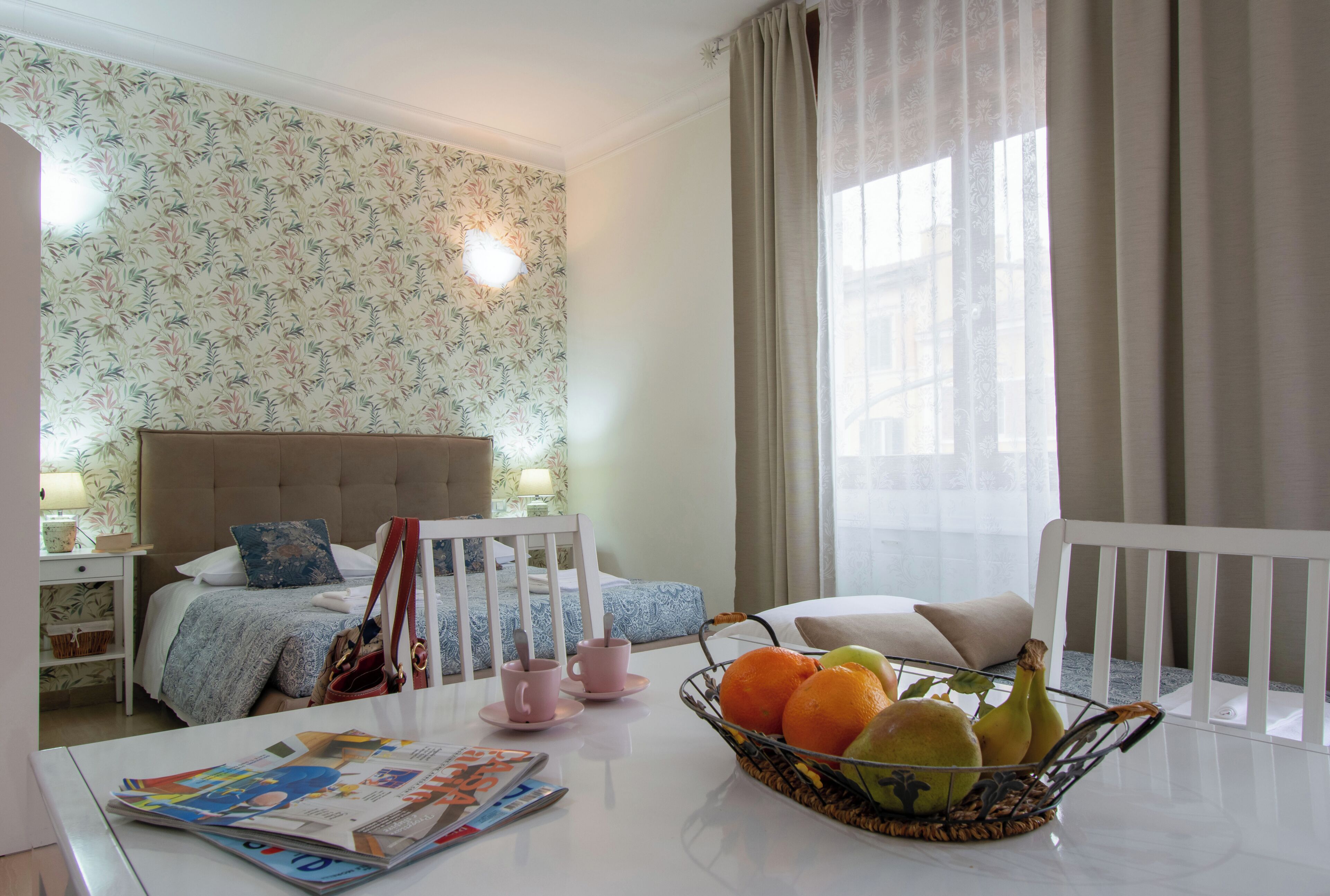 Quarto de hotel com uma cama, uma mesa com uma cesta de frutas, uma revista e um abajur.