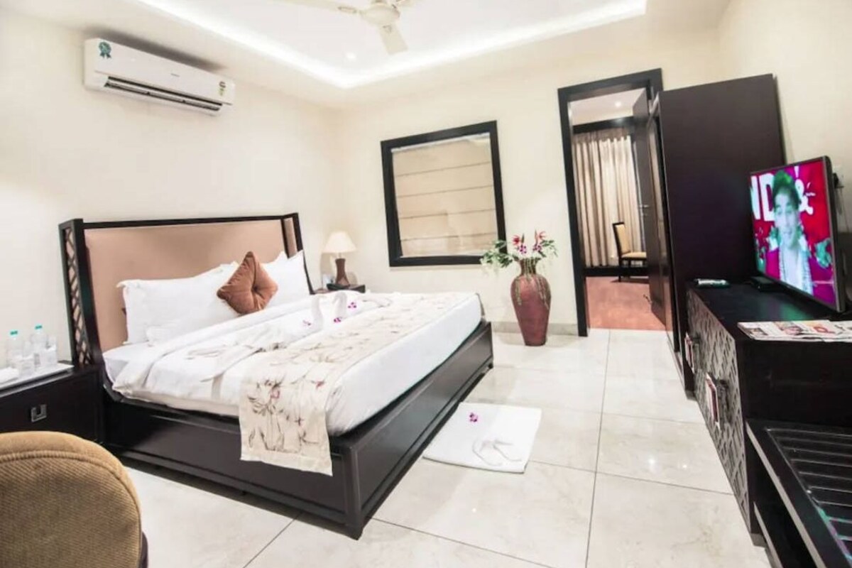 Quarto de hotel com cama, televisão, ar condicionado, um vaso com flores e uma porta que dá para outro quarto.