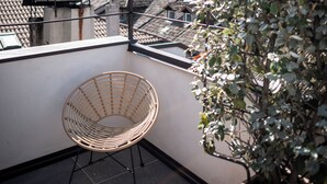 Uma varanda com uma cadeira de vime, um vaso de planta e vista para edifícios e montanhas.