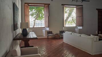 Uma sala de estar ampla, com piso de azulejo, um sofá, uma mesa com um vaso, e duas janelas com venezianas de madeira.