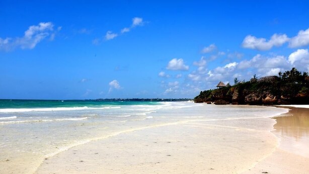 Uma praia com águas azul-claras, areia dourada e um afloramento rochoso com vegetação.