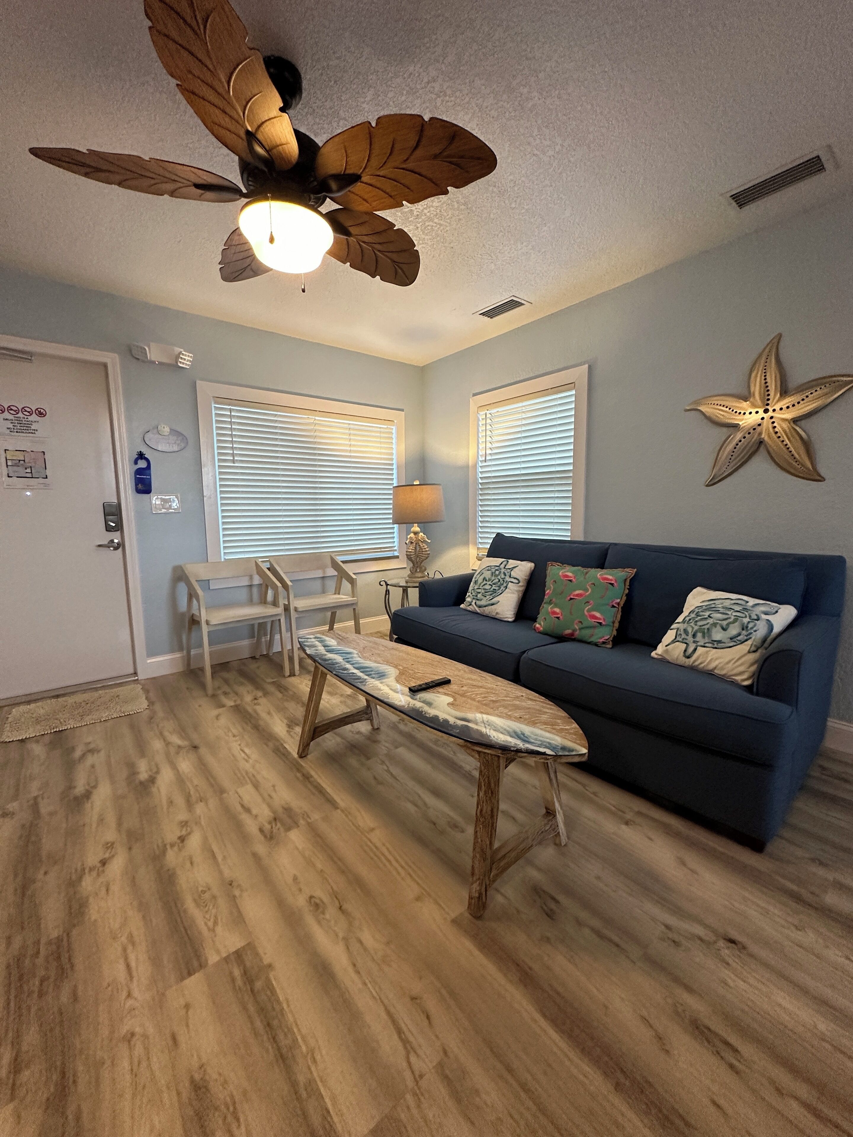 Uma sala de estar com piso de madeira, um sofá azul com almofadas decorativas, uma mesa de centro de madeira e um ventilador de teto com lâminas de madeira.