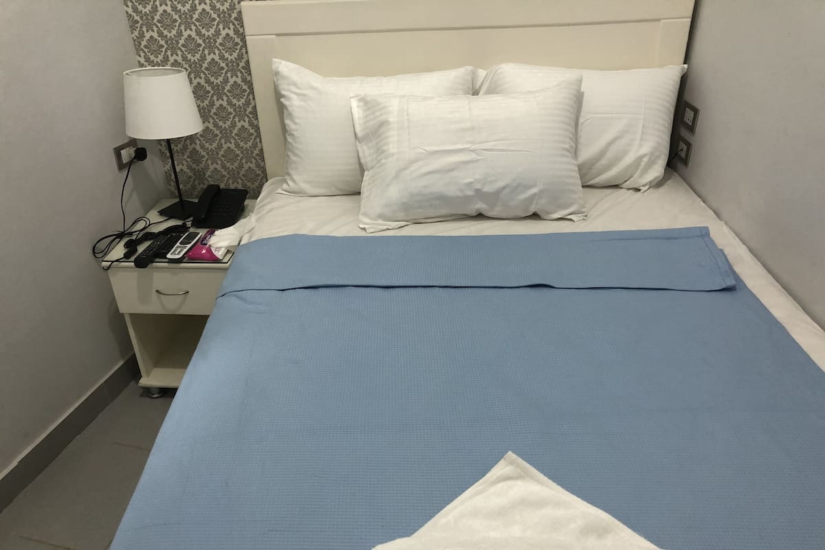 Uma cama bem arrumada com almofadas brancas e um cobreleite azul, uma mesa de cabeceira com abajur e telefone, e uma parede estampada.