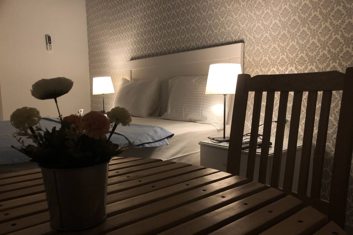 Quarto de hotel com uma cama, duas luminárias de cabeceira, uma cadeira de madeira e um vaso de plantas com flores.