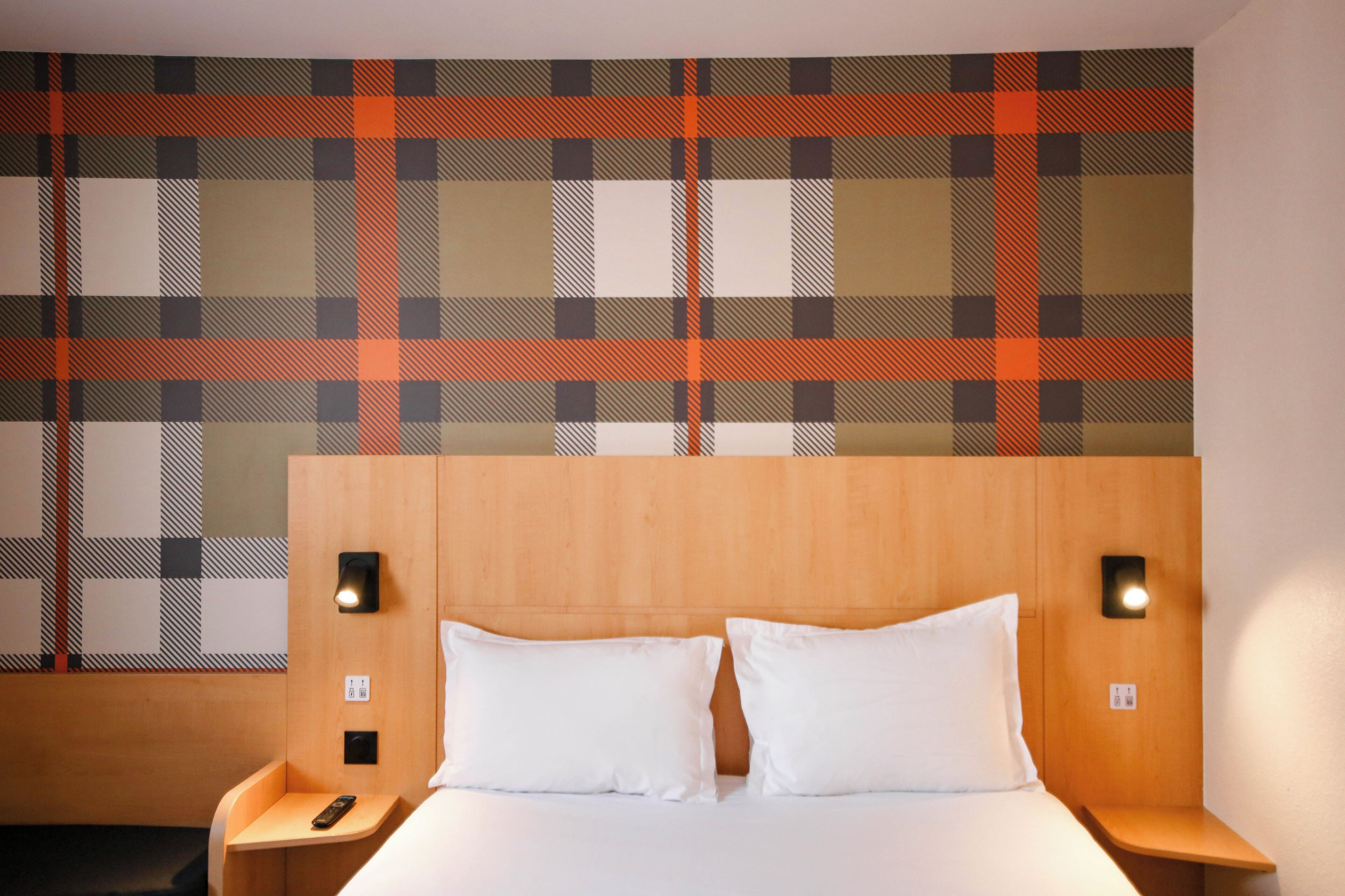 Quarto de hotel com padrão quadriculado na parede, cabeceira de madeira, duas almofadas brancas e luminárias laterais.