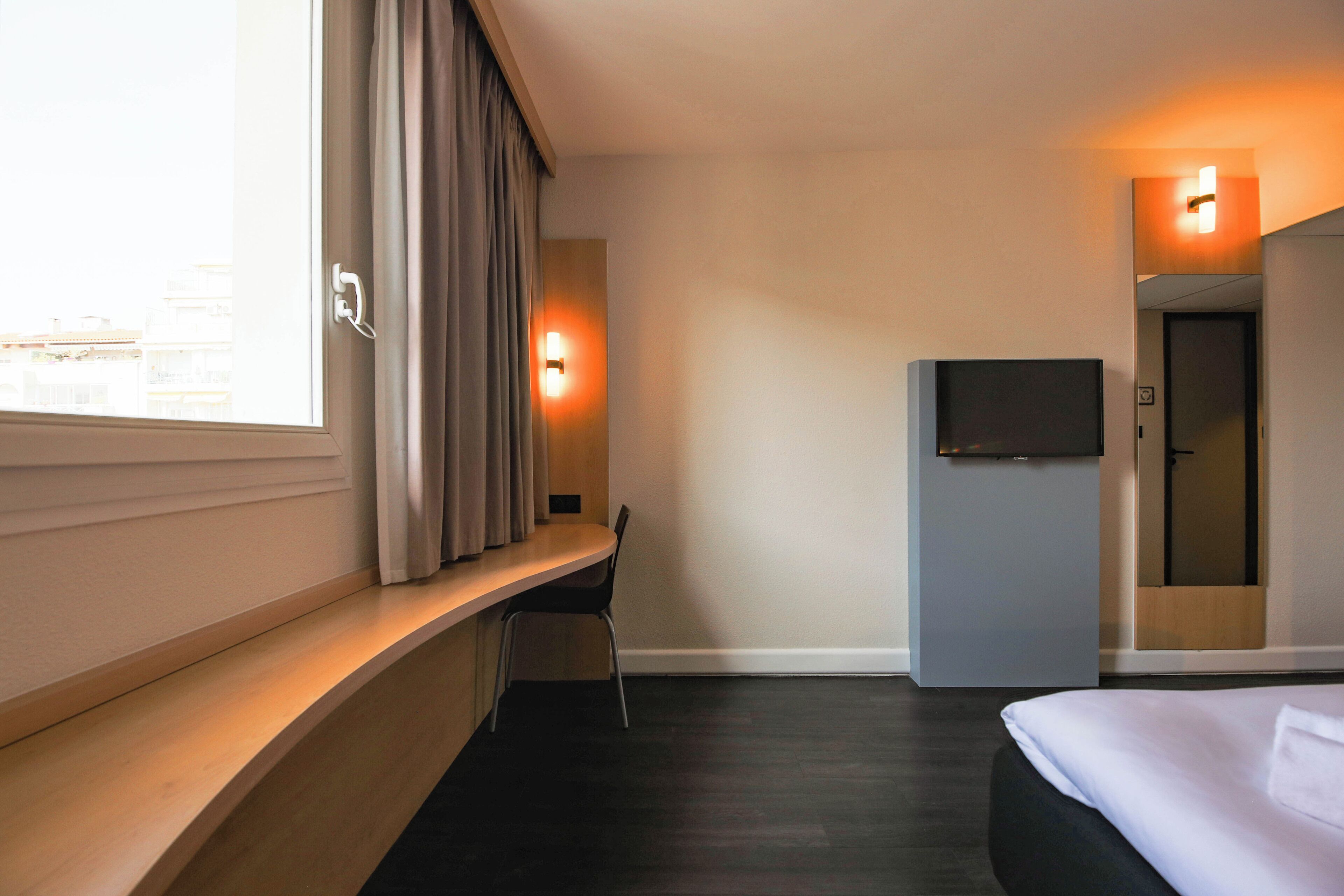 Quarto de hotel com cama, escrivaninha, cadeira, televisão e janela com cortinas.
