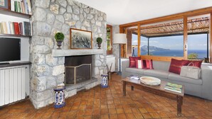 Uma sala de estar com lareira de pedra, televisão, sofá com almofadas e vista para o mar.