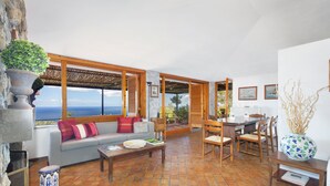 Ampla sala de estar com grande janela vista para o oceano, mesa de jantar de madeira e um espaço de estar acolhedor.