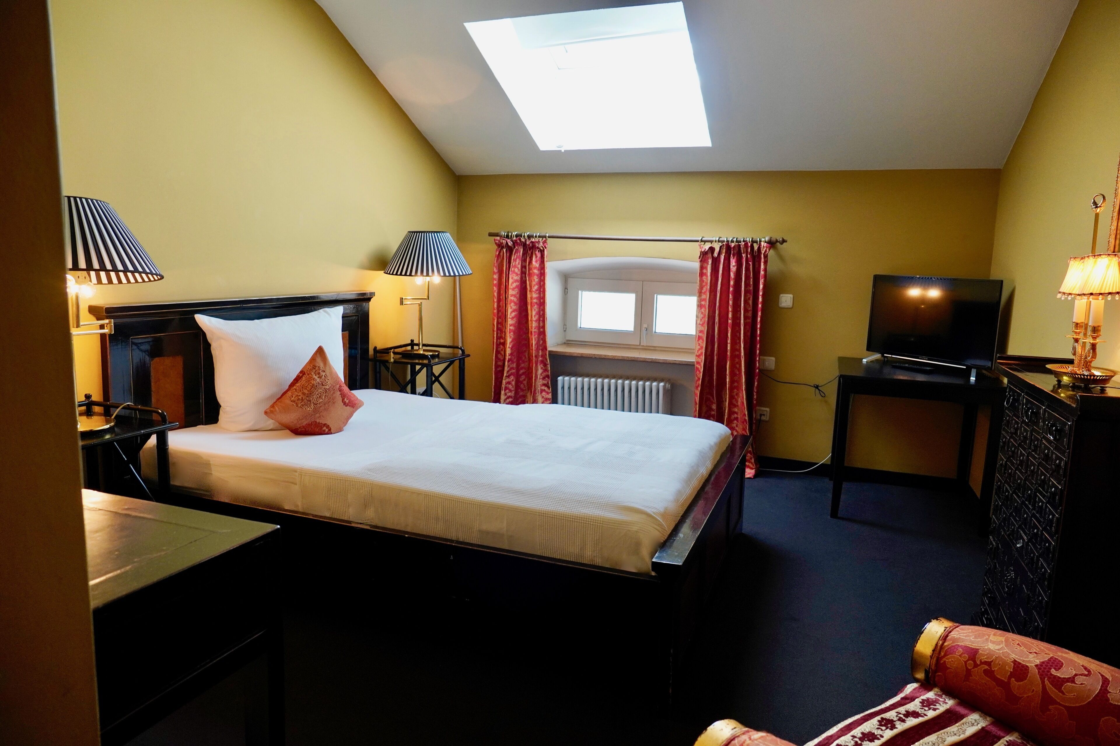 Quarto de hotel com cama, escrivaninha, televisão e janela com cortinas.