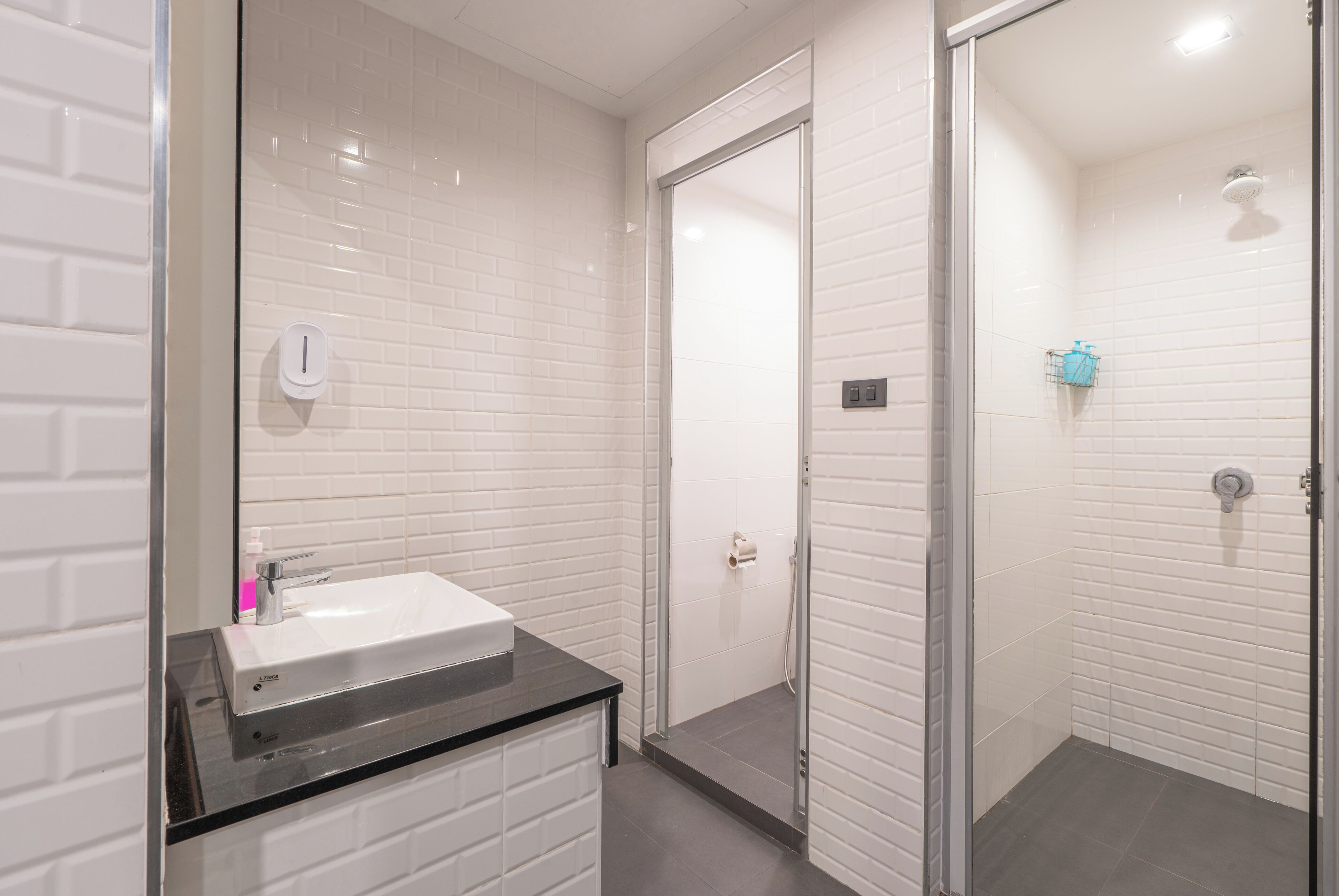 Casa de banho com parede revestida a azulejos brancos, bancada preta, cabine de chuveiro em vidro e lavatório branco.
