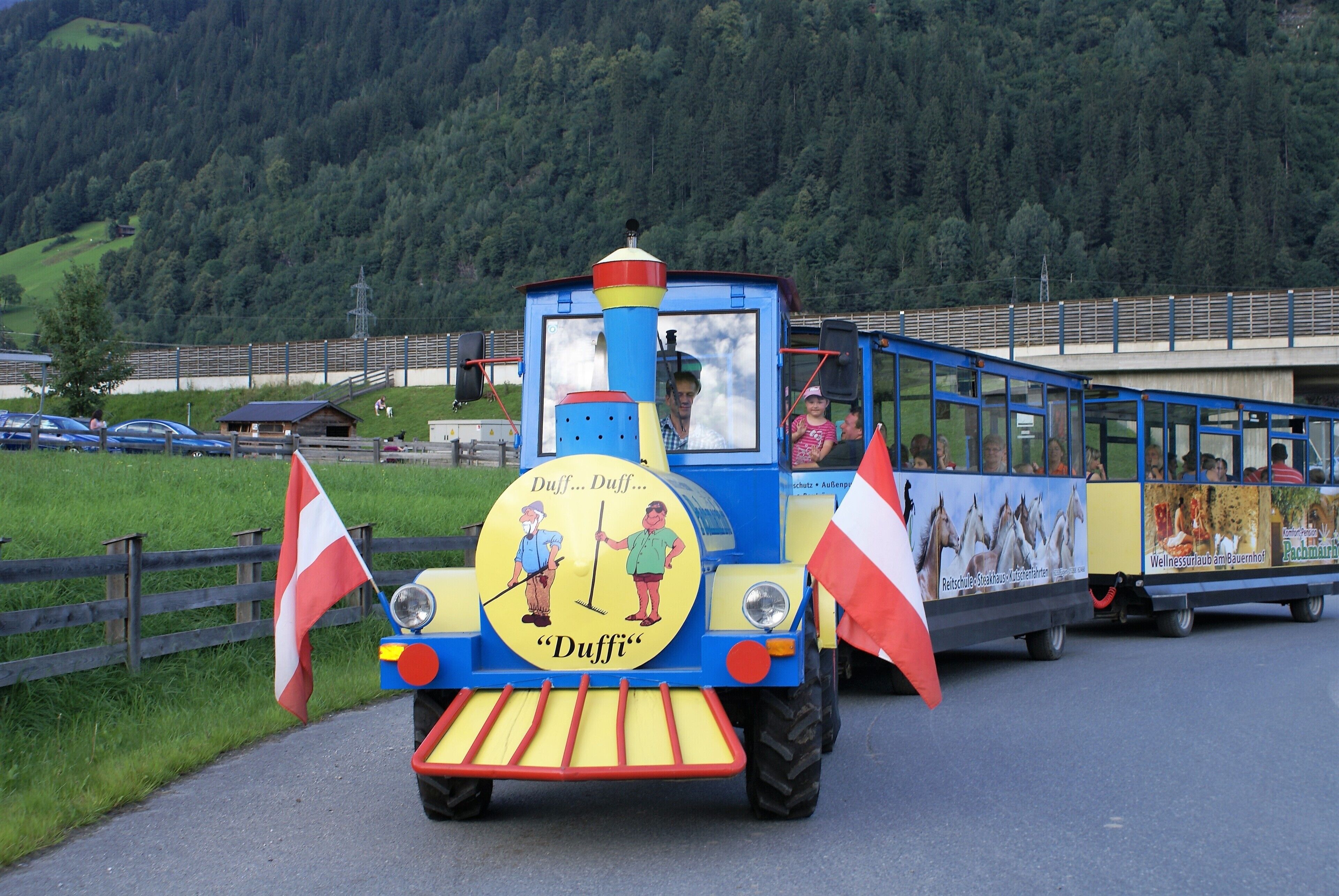 Um trem colorido com a marcação 'Duffi' e uma bandeira suíça, seguindo por uma estrada com uma paisagem arborizada ao fundo.