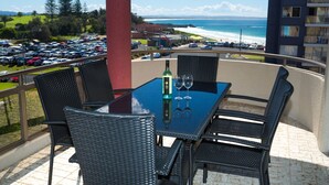 Varanda com mesa e cadeiras, vista para um estacionamento e praia.