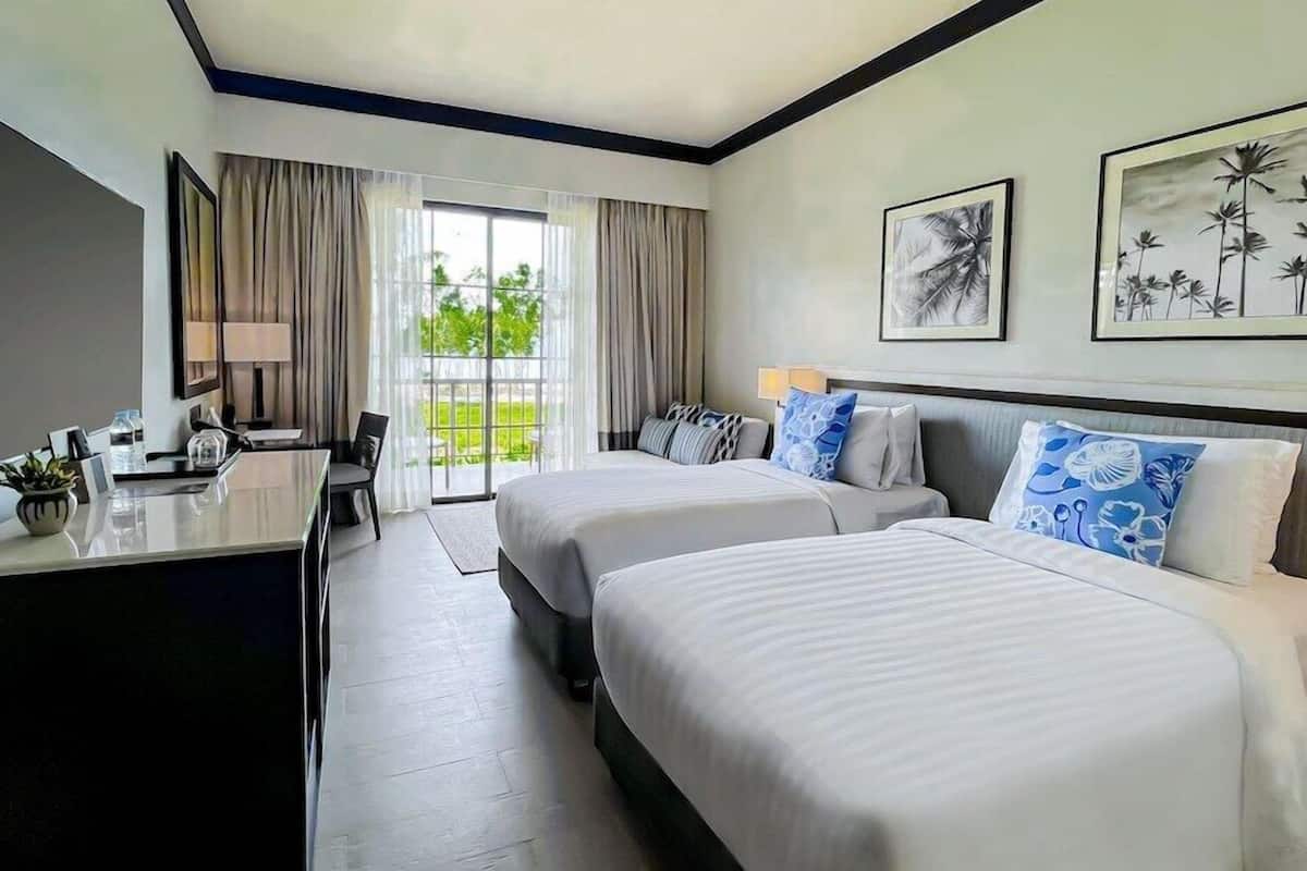 Quarto de hotel com duas camas, uma escrivaninha, uma cadeira e uma janela grande com cortinas.