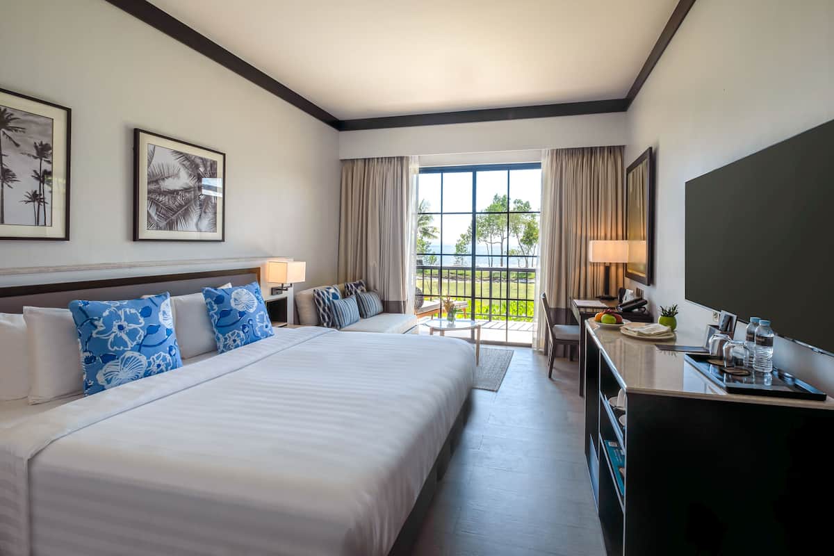 Quarto de hotel com uma cama grande, uma escrivaninha com computador, televisão e varanda com vista para o mar.