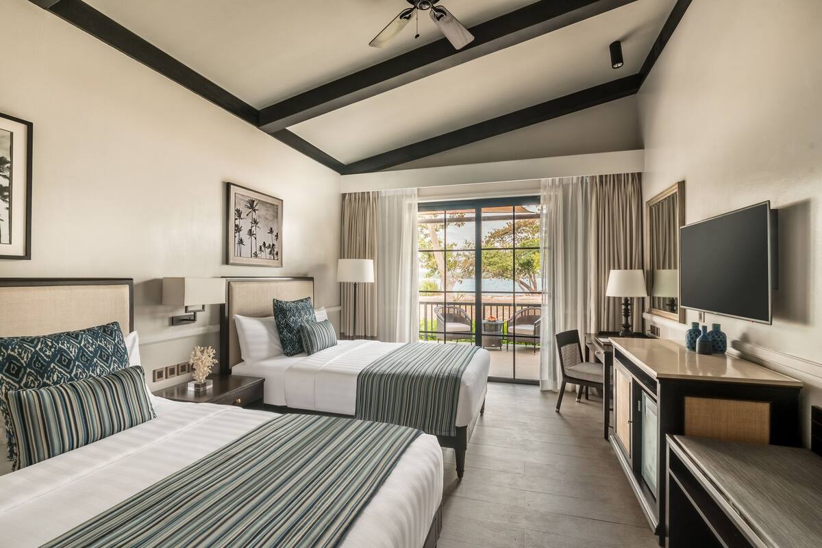 Quarto de hotel com duas camas, uma escrivaninha, televisão e varanda com cortinas.